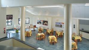 Uma ampla sala de jantar com várias mesas e cadeiras, um extintor de incêndio e vista para o exterior através de grandes janelas.