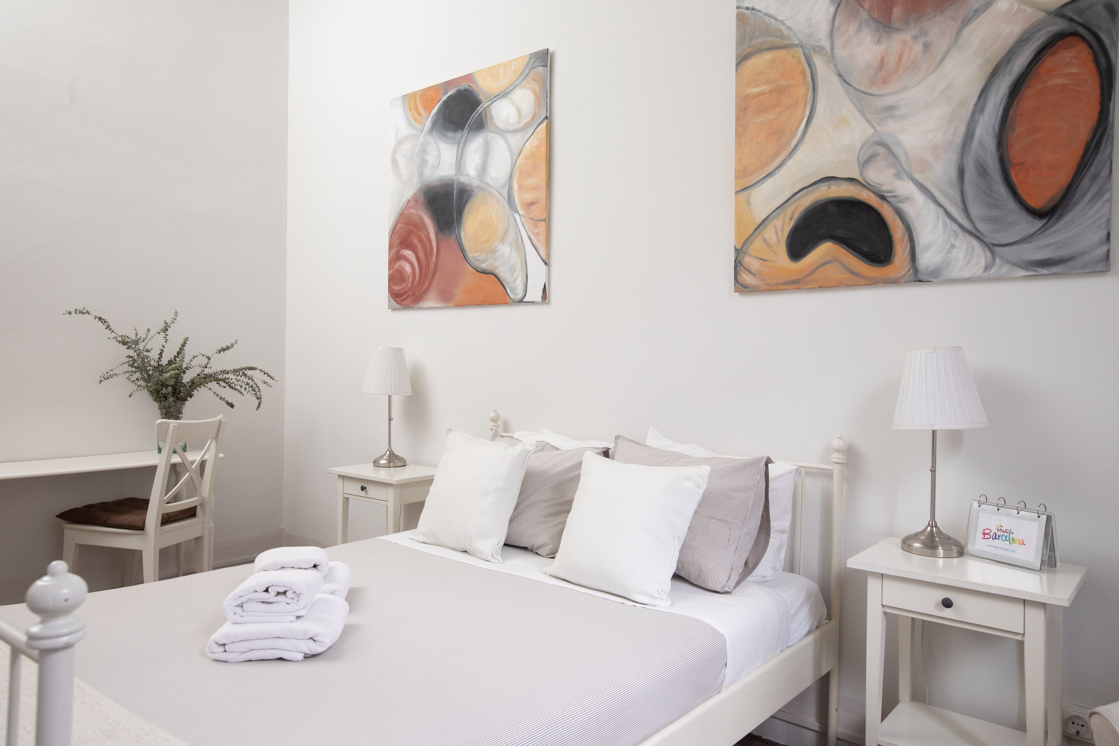 Quarto de hotel com cama bem arrumada, escrivaninha e cadeira. Há duas pinturas na parede.
