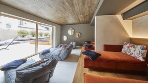 Quarto moderno com uma janela grande, teto de madeira e área de estar com sofás laranja e cinza.