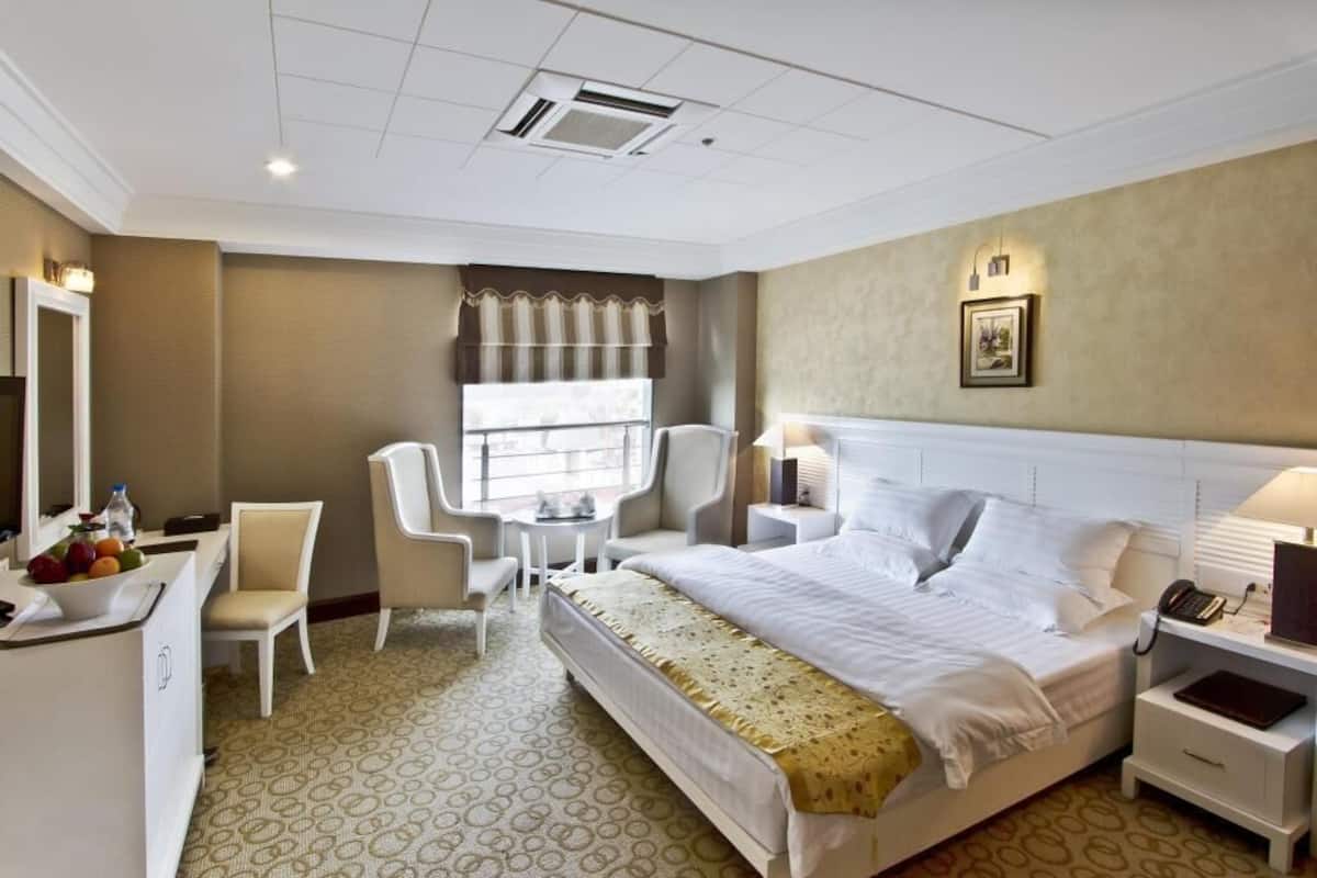 Quarto de hotel com cama, mesas de cabeceira, escrivaninha com cadeiras, janela com cortinas e um quadro na parede.