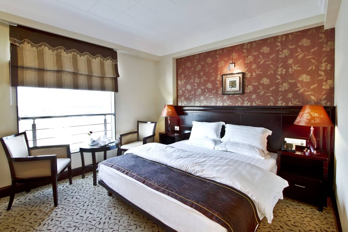 Quarto de hotel com uma cama grande, duas mesas de cabeceira com luminárias, uma cadeira, uma mesinha e uma janela com cortinas.