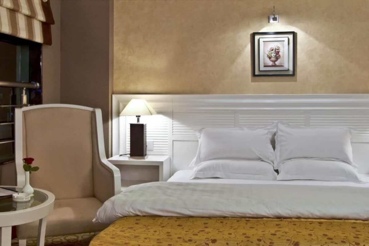 Quarto de hotel com cama, mesa de cabeceira com abajur, cadeira e uma pequena mesa com um vaso.