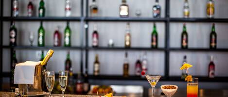 Uma área de bar com bancada de granito, diversos frascos em prateleiras e três copos com bebidas diferentes.