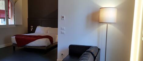 Quarto de hotel com cama, mesa de cabeceira, abajur e sofá.