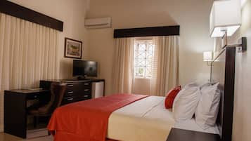 Quarto de hotel com cama, escrivaninha, cadeira, televisão e janela com cortinas.