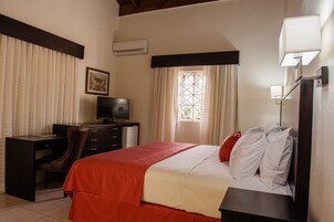 Quarto de hotel com cama, escrivaninha, cadeira, televisão e janela com cortinas.