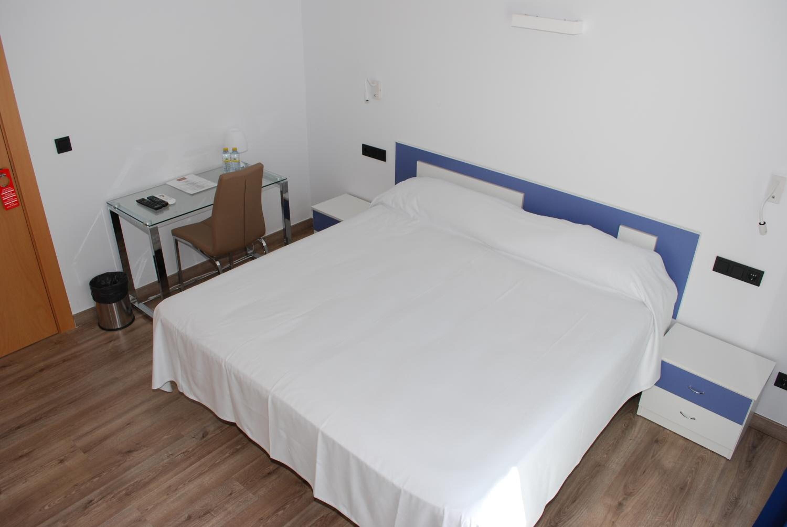 Quarto de hotel com uma cama de solteiro, uma pequena escrivaninha, uma cadeira, um cesto de lixo e uma mesa de cabeceira com abajur.