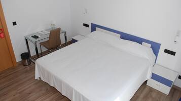 Quarto de hotel com uma cama de solteiro, uma pequena escrivaninha, uma cadeira, um cesto de lixo e uma mesa de cabeceira com abajur.