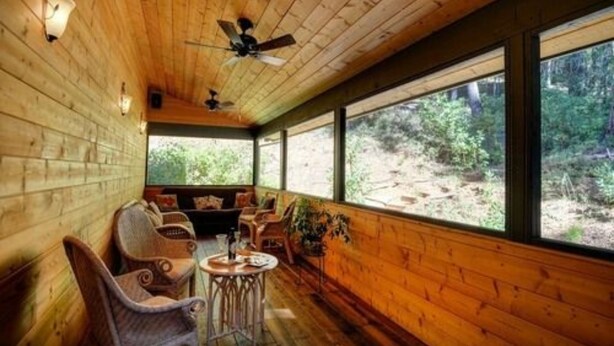 Interior de uma cabana de madeira com ventilador de teto, janelas amplas, área de estar com cadeiras e uma mesa, e vista para a floresta.