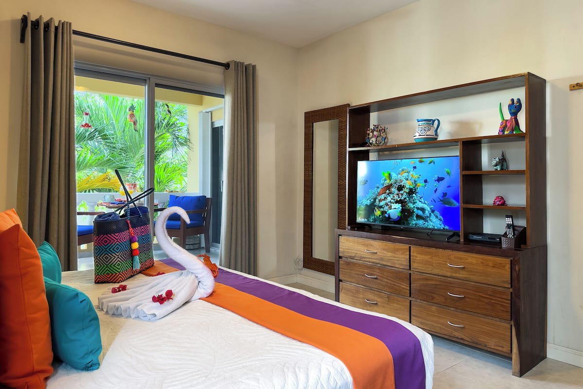 Quarto de hotel com cama, televisão, cómoda de madeira e vista para o exterior com vegetação.
