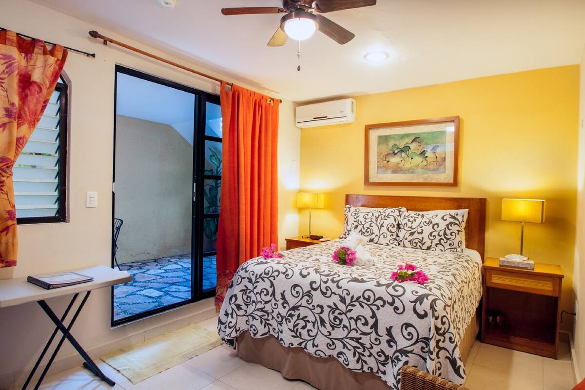 Um quarto com cama, duas mesas de cabeceira, um ventilador de teto, aparelho de ar condicionado, janela com cortinas e um quadro na parede.