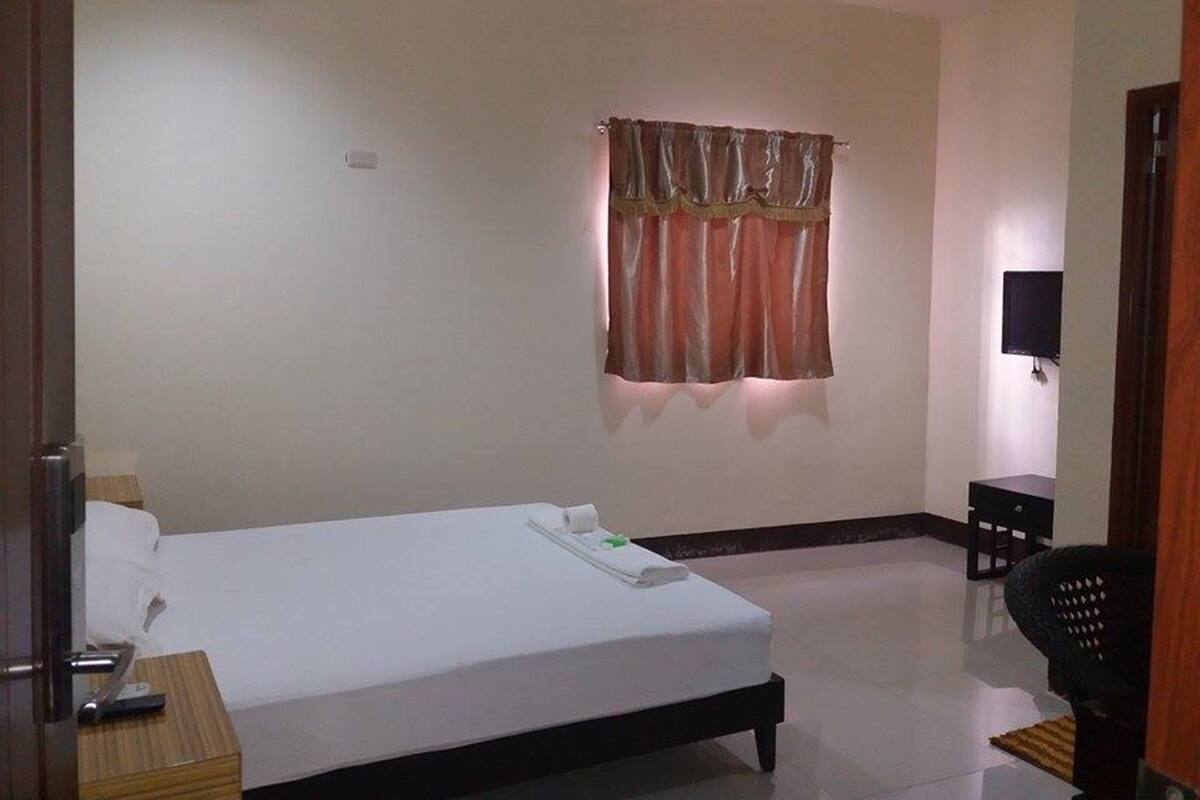 Quarto de hotel com cama, escrivaninha, cadeira, televisão e janela com cortinas.