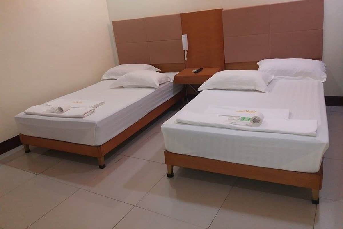 Duas camas de solteiro com cabeceiras de madeira e roupa de cama branca, uma mesa de cabeceira com controle remoto e luminária fixada na parede.