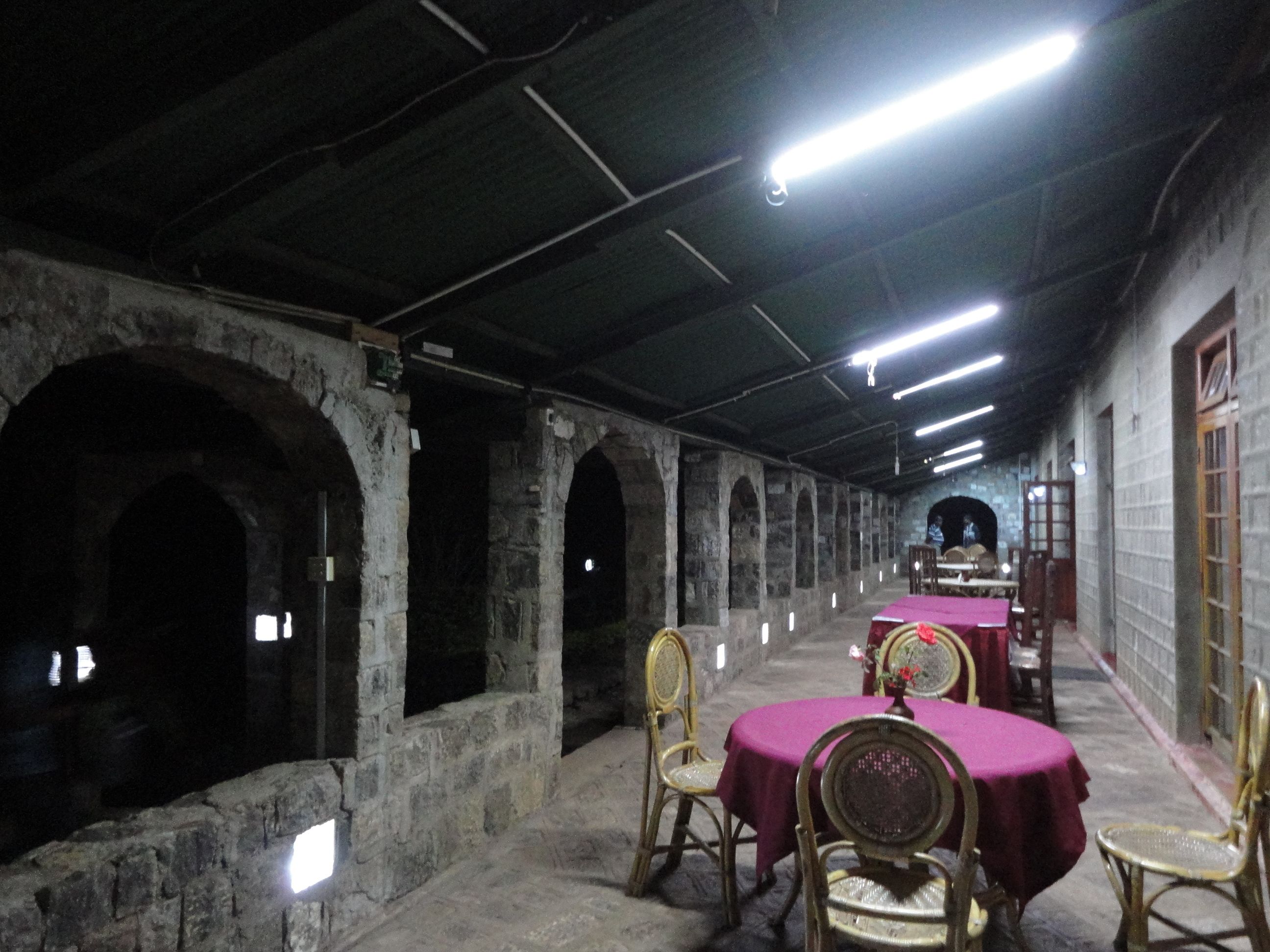 Um corredor mal iluminado com arcos de pedra e mesas cobertas por toalhas vermelhas.