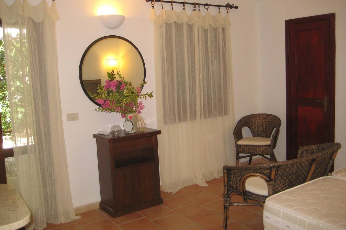 Um quarto com uma cama, uma cadeira, um armário de madeira, um espelho e uma janela com cortinas.