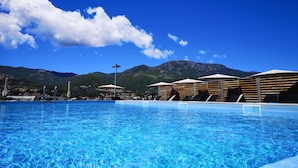 Piscina azul cristalina com espreguiçadeiras e guarda-sóis, rodeada por montanhas e céu limpo.