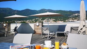 Mesa de café da manhã com croissants, torradas, frutas e bebidas, com vista para uma paisagem montanhosa.