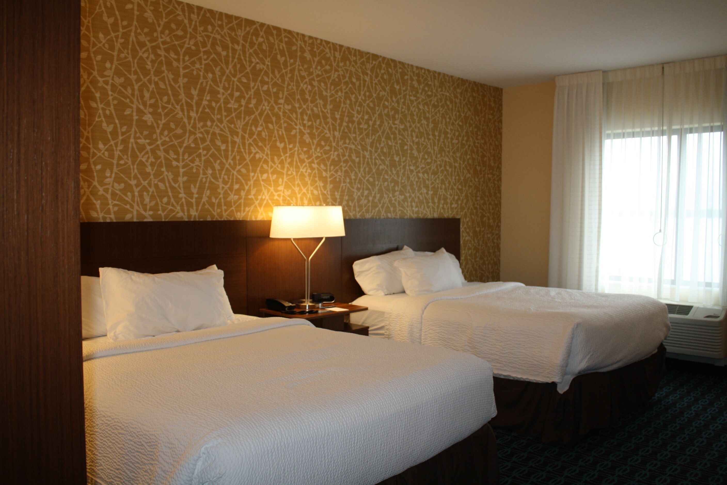Quarto de hotel com duas camas, uma mesinha de cabeceira com abajur, papel de parede estampado e uma janela com cortinas.
