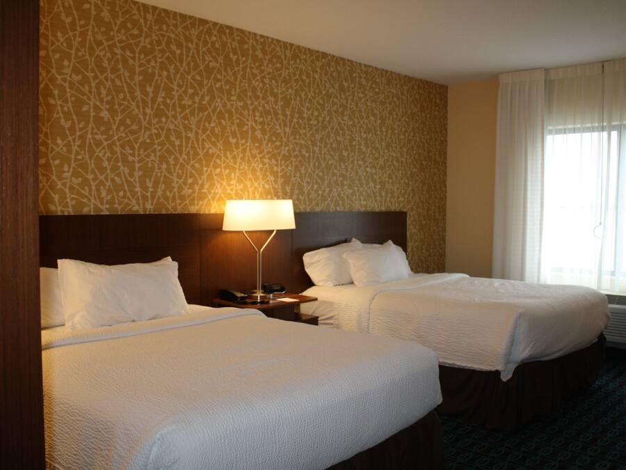 Quarto de hotel com duas camas, uma mesinha de cabeceira com abajur, papel de parede estampado e uma janela com cortinas.