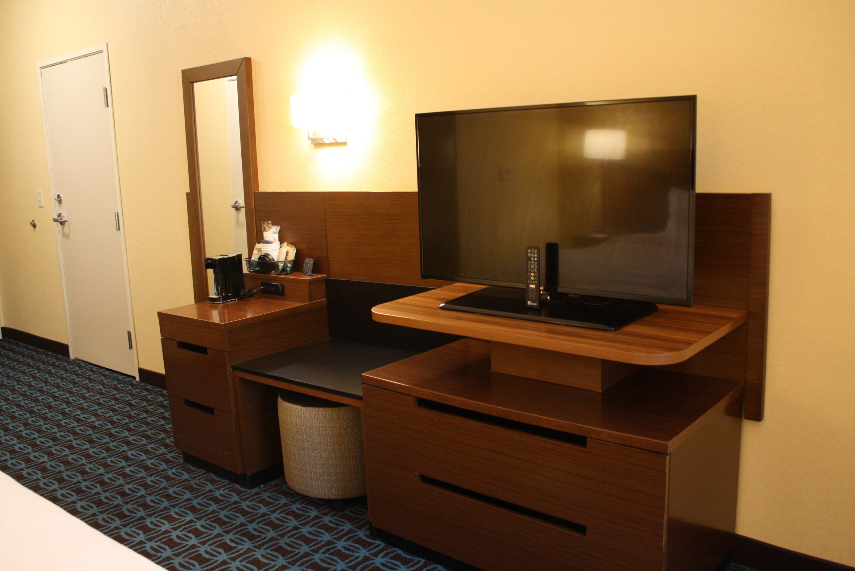 Quarto de hotel com escrivaninha de madeira, televisão de tela plana, espelho e lixeira.