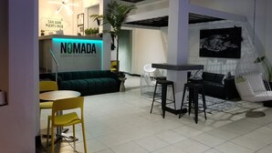 Interior de um hostel urbano moderno com um sofá verde, cadeiras amarelas, uma mesa de jantar e um espaço de bar.