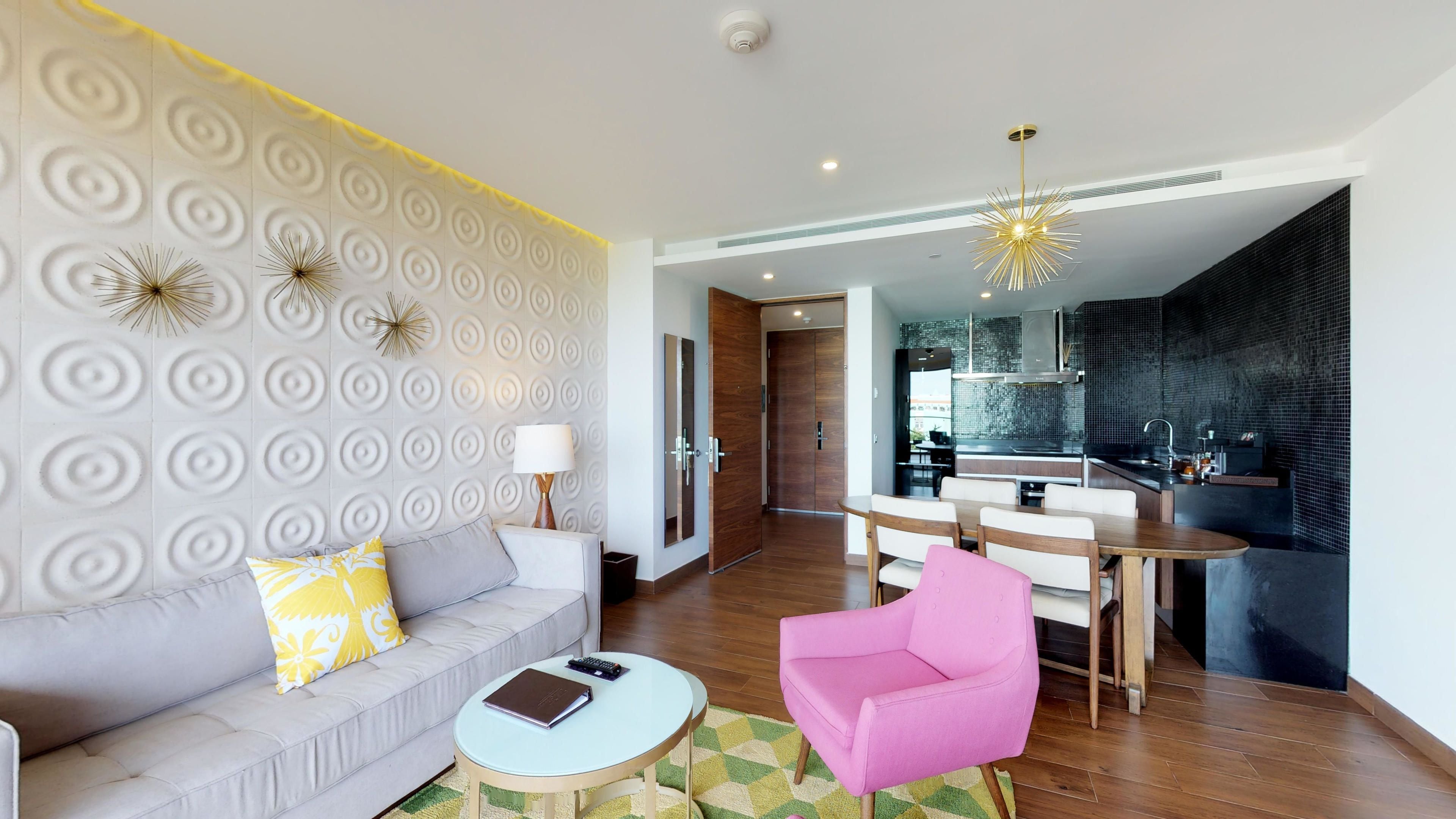 Uma sala de estar moderna com um sofá, uma poltrona rosa e uma mesa de centro. Uma cozinha é visível ao fundo, com um painel de fundo preto e eletrodomésticos de aço inoxidável.