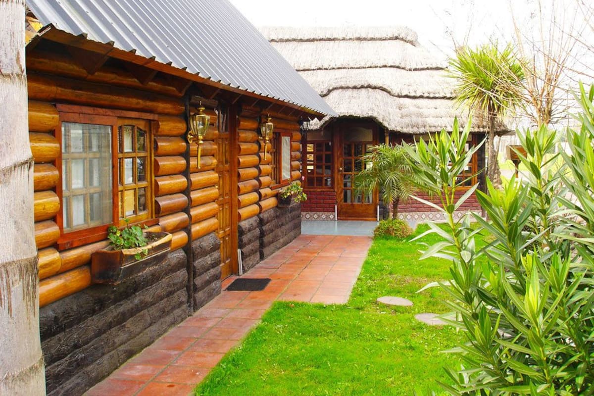 Uma cabana de tronco com telhado de palha, fachada de madeira e um pequeno jardim com plantas e um caminho.