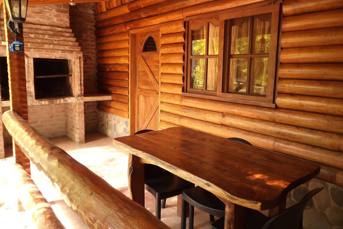 Uma cabana de madeira com lareira de tijolos, uma mesa de jantar e uma janela com cortinas.