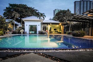 Piscina com deck ladrilhado, rodeada por plantas em vasos e um pequeno edifício com porta de vidro.
