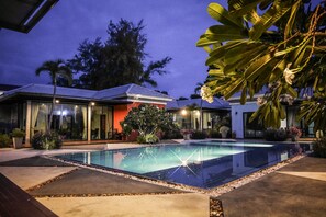 Uma casa moderna com piscina, rodeada por plantas tropicais e um céu claro ao anoitecer.