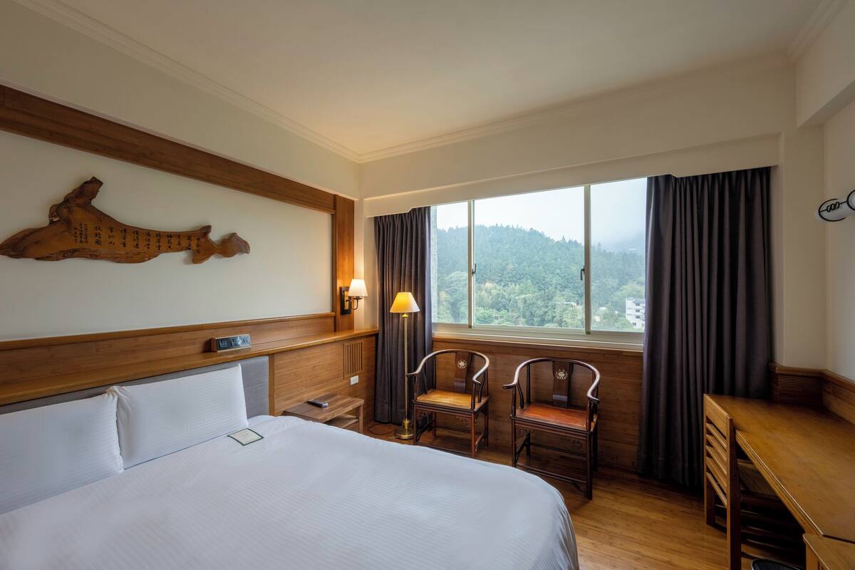 Quarto de hotel com uma cama grande, duas cadeiras de madeira, uma mesa pequena, um criado-mudo, uma luminária e vista para uma floresta pela janela.