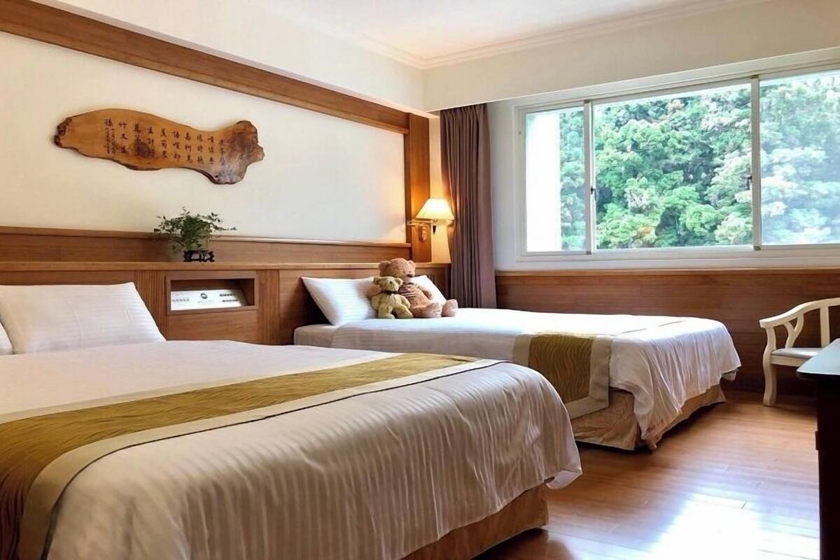Quarto de hotel com duas camas, cabeceira de madeira, mesa de cabeceira com abajur, janela com vista para árvores e um painel de madeira decorativo na parede.