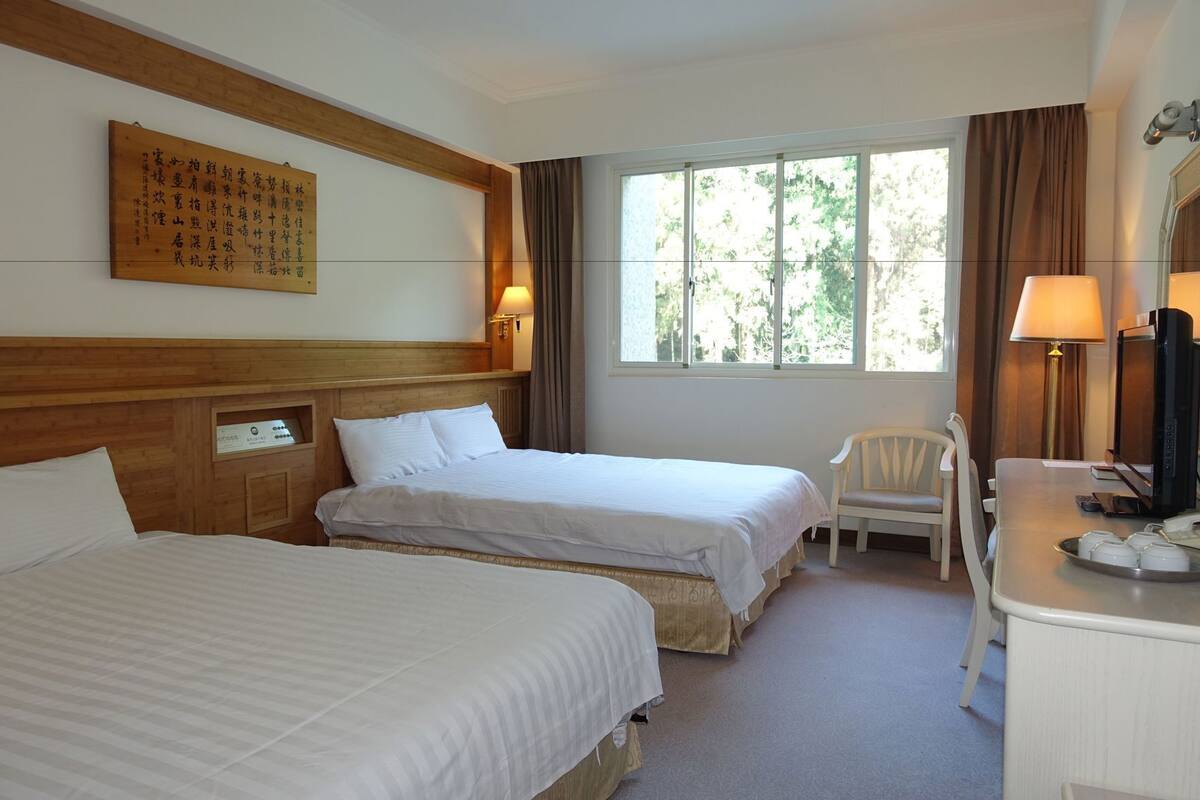 Quarto de hotel com duas camas, uma mesa de trabalho com cadeira, televisão e janela com cortinas.