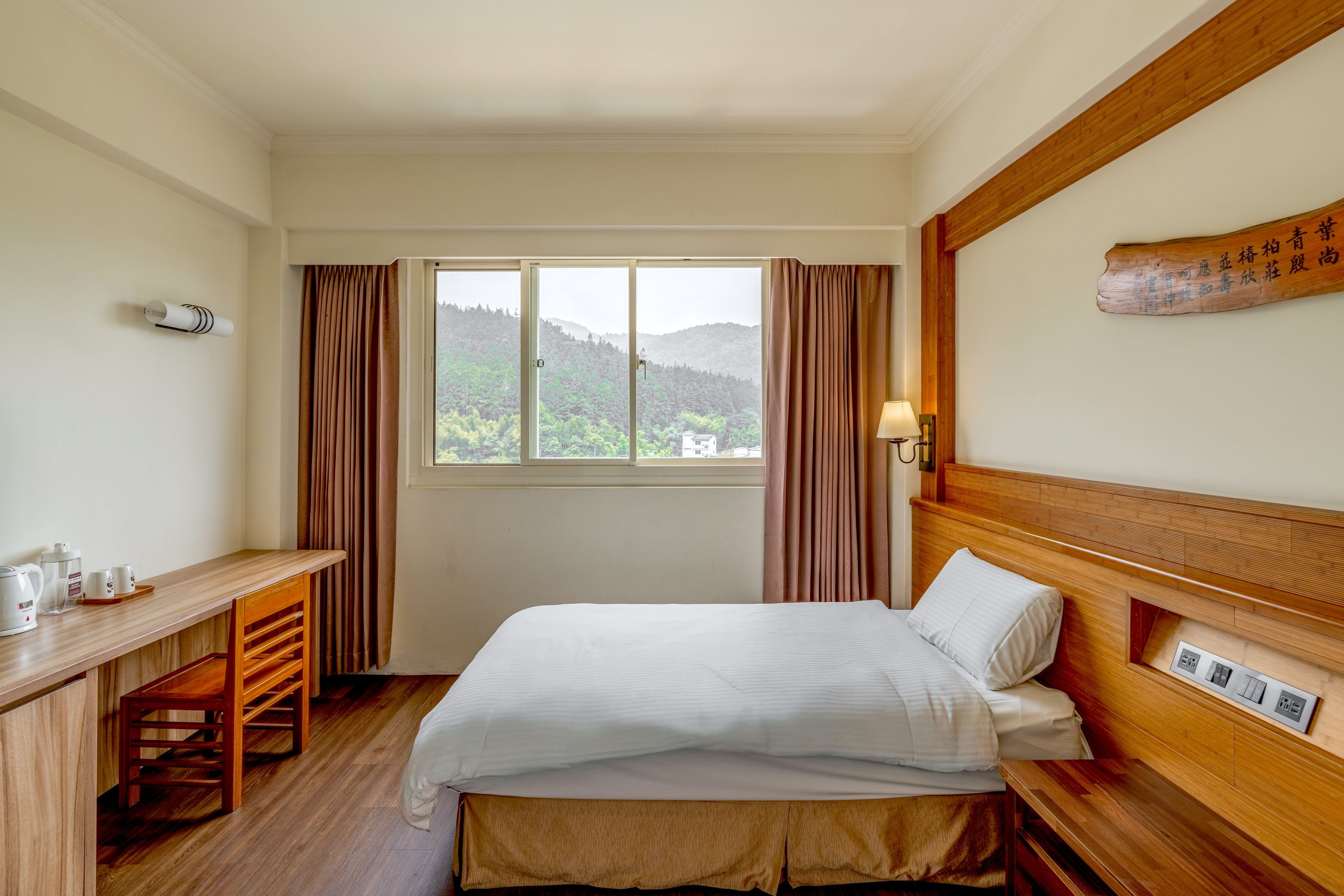 Quarto de hotel com cama, escrivaninha, cadeira, janela com cortinas e vista para uma área arborizada.