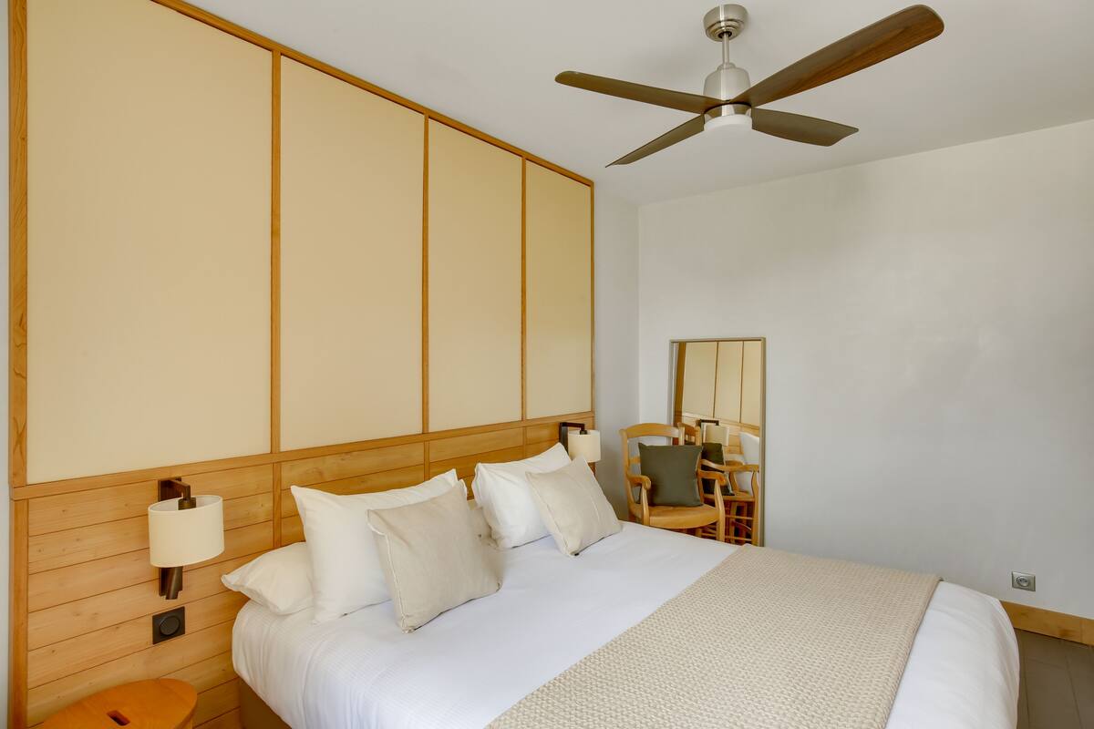 Quarto de hotel com uma cama grande, um ventilador de teto, uma mesa com cadeira e uma luminária na parede.
