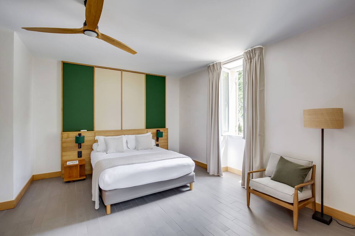 Quarto de hotel moderno com cama grande, ventoinha de teto em madeira, mesa de cabeceira com luminária, uma cadeira e vista para o exterior pela janela.