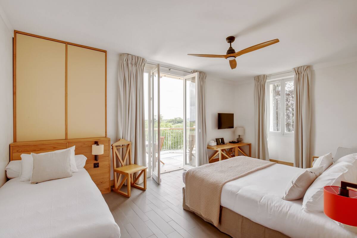Quarto de hotel com duas camas, uma mesa, uma cadeira, uma televisão e uma varanda com cortinas.