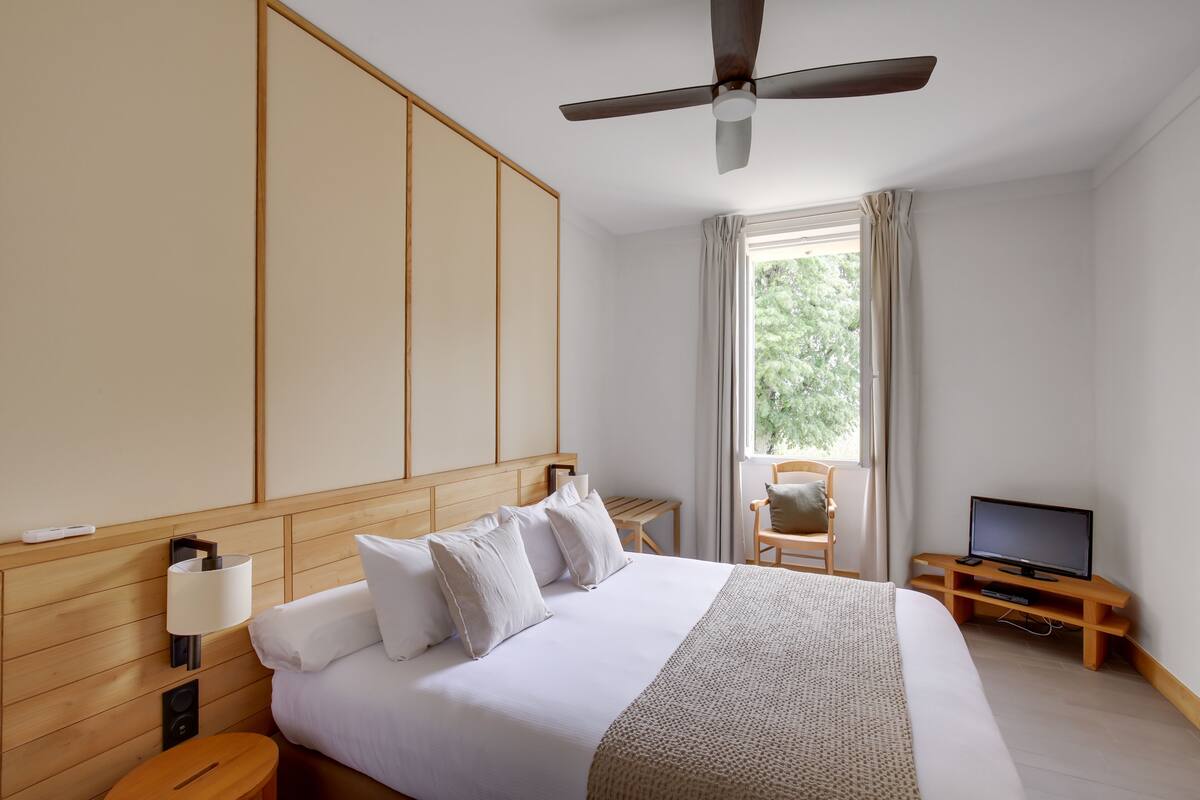 Quarto de hotel com cama grande, ventilador de teto, televisão e janela com cortinas.