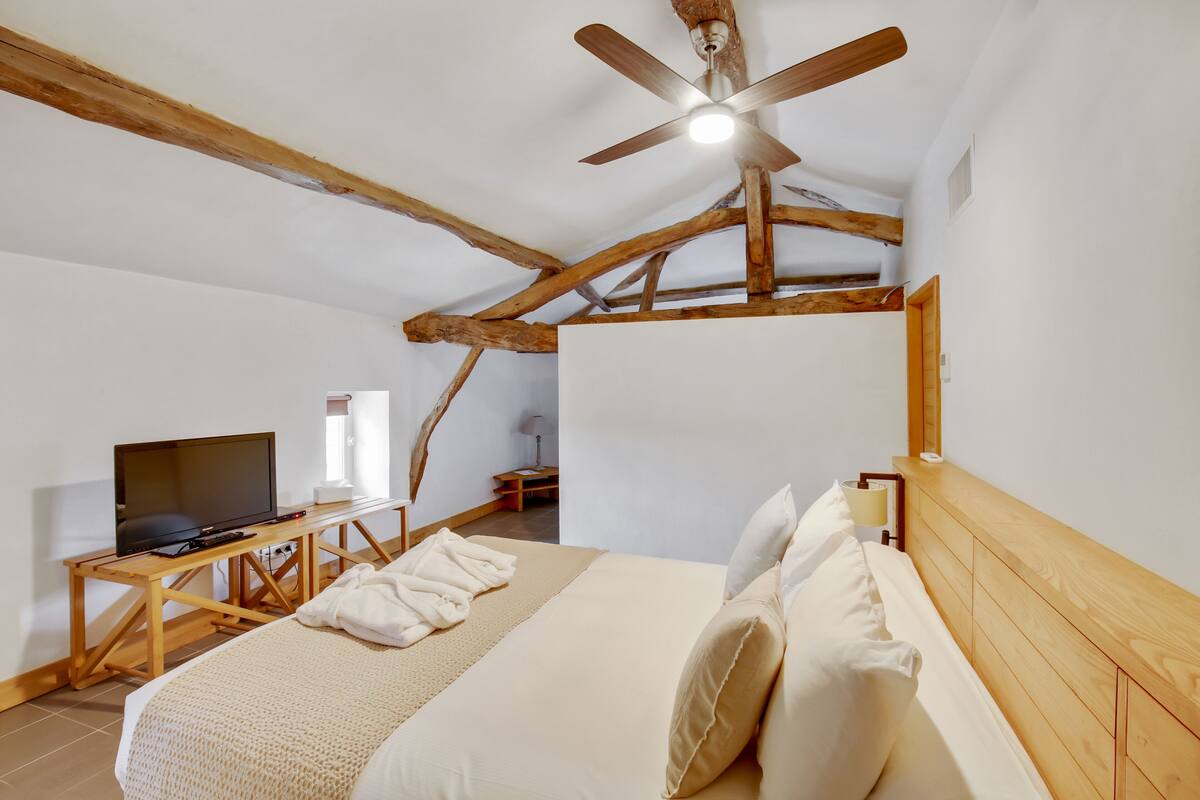 Quarto com teto de madeira, cama com roupa de cama branca, televisão sobre uma escrivaninha de madeira e um ventilador de teto.