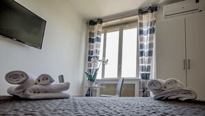 Quarto de hotel com uma cama, televisão fixada na parede, janela com cortinas, planta em vaso e um porta-toalhas.