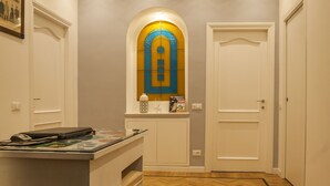 Um corredor com piso em padrão herringbone, uma recepcionista branca, um quadro na parede e uma porta com um pequeno vitrô.