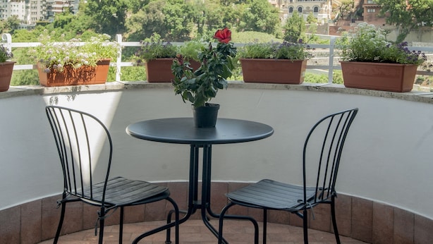 Mesa redonda com duas cadeiras numa varanda com plantas em vasos e vista para edifícios.