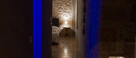 Corredor estreito com parede de pedra de um lado e porta de madeira do outro. Uma luminária é visível ao longe, e o corredor dá para um quarto com cama e mesa.