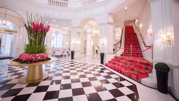 Um hall imponente com piso xadrez, escada com tapete vermelho e um grande arranjo de flores.