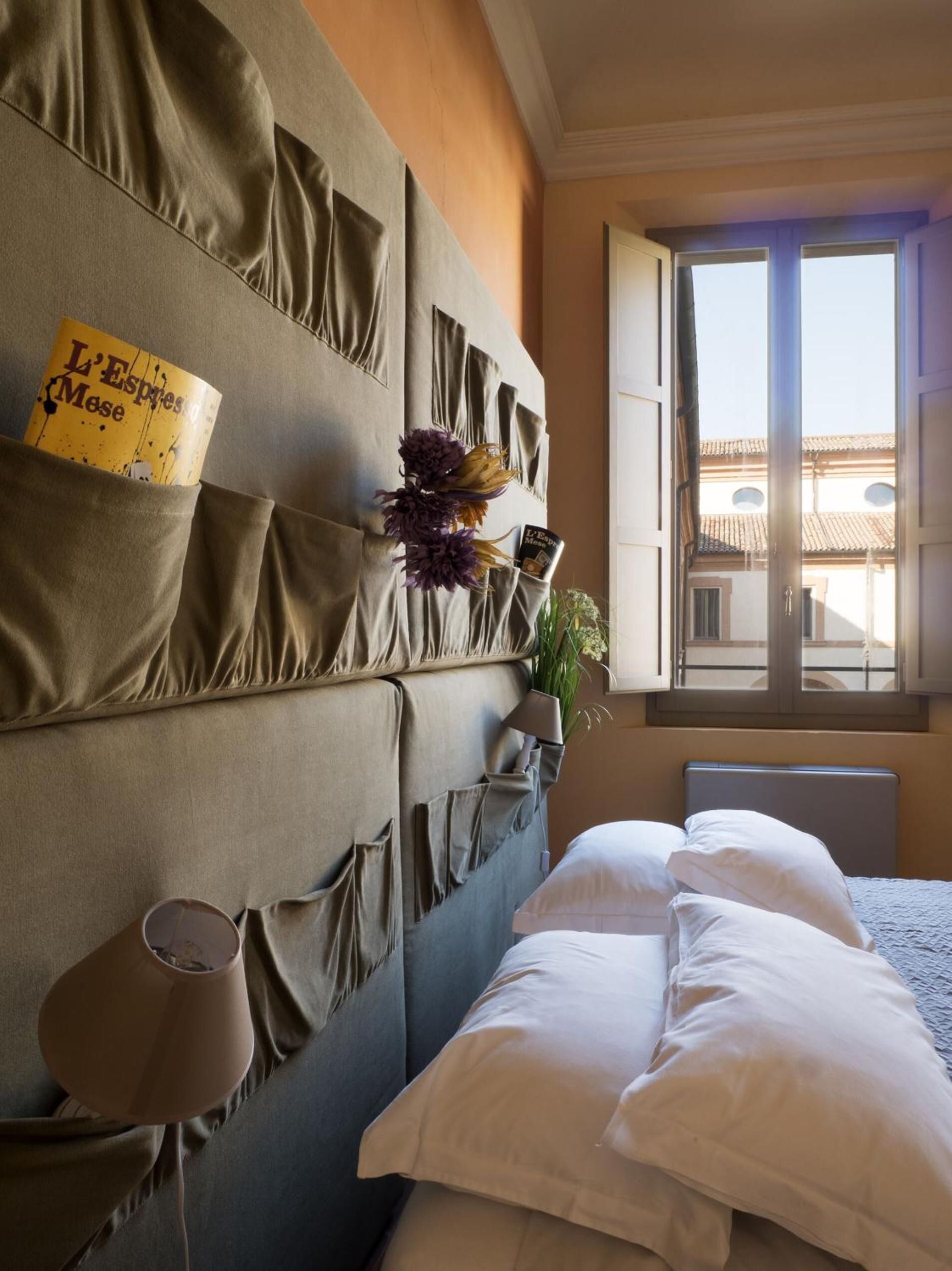Quarto de hotel com uma cabeceira grande, uma janela com vista para edifícios e uma luminária na mesa lateral.