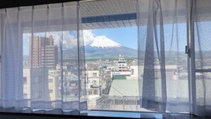Vista pela janela, com cortinas leves, mostrando um horizonte urbano com edifícios e uma montanha ao fundo.