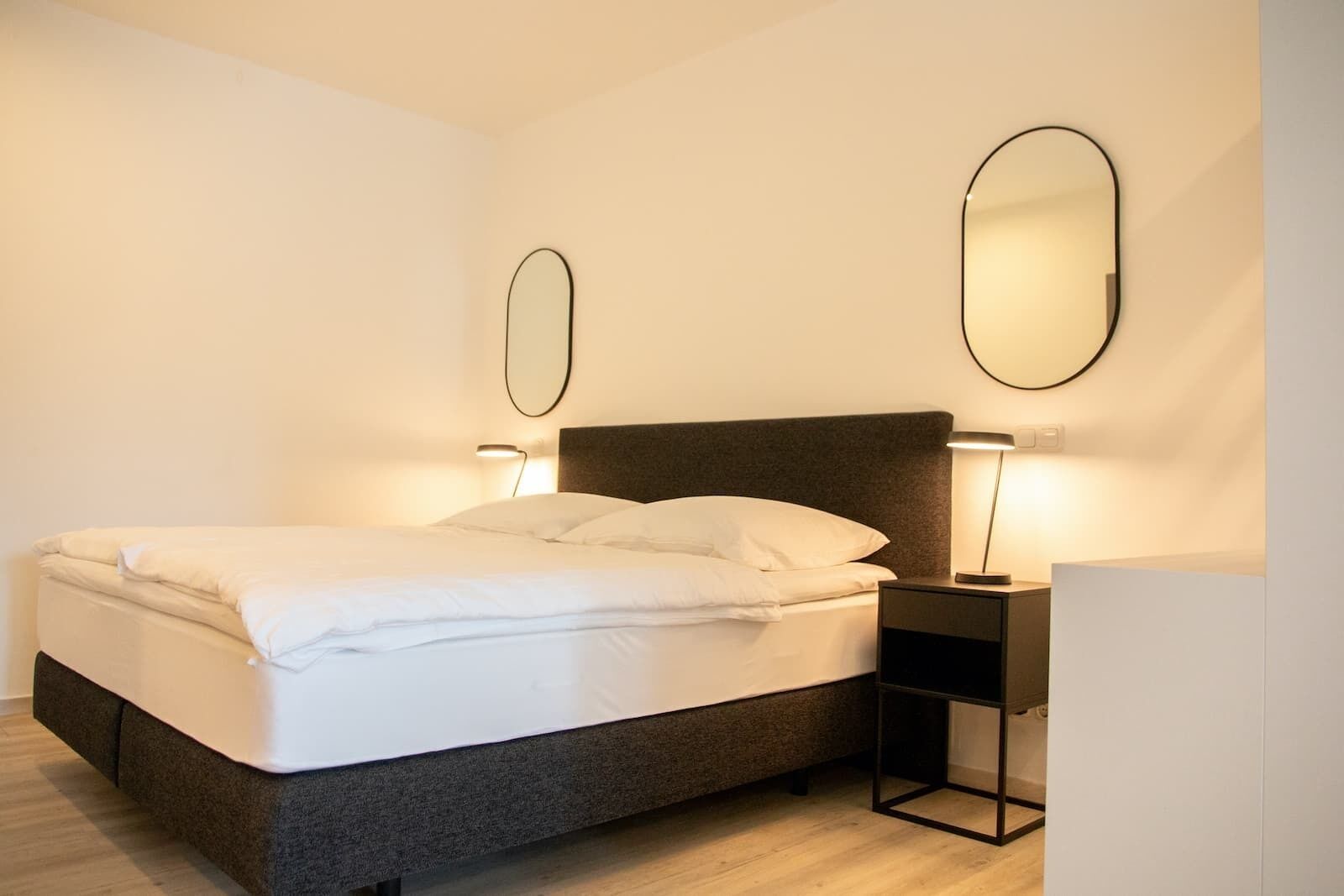 Quarto moderno com cabeceira cinza escura, roupa de cama branca e duas espelhos fixados na parede.