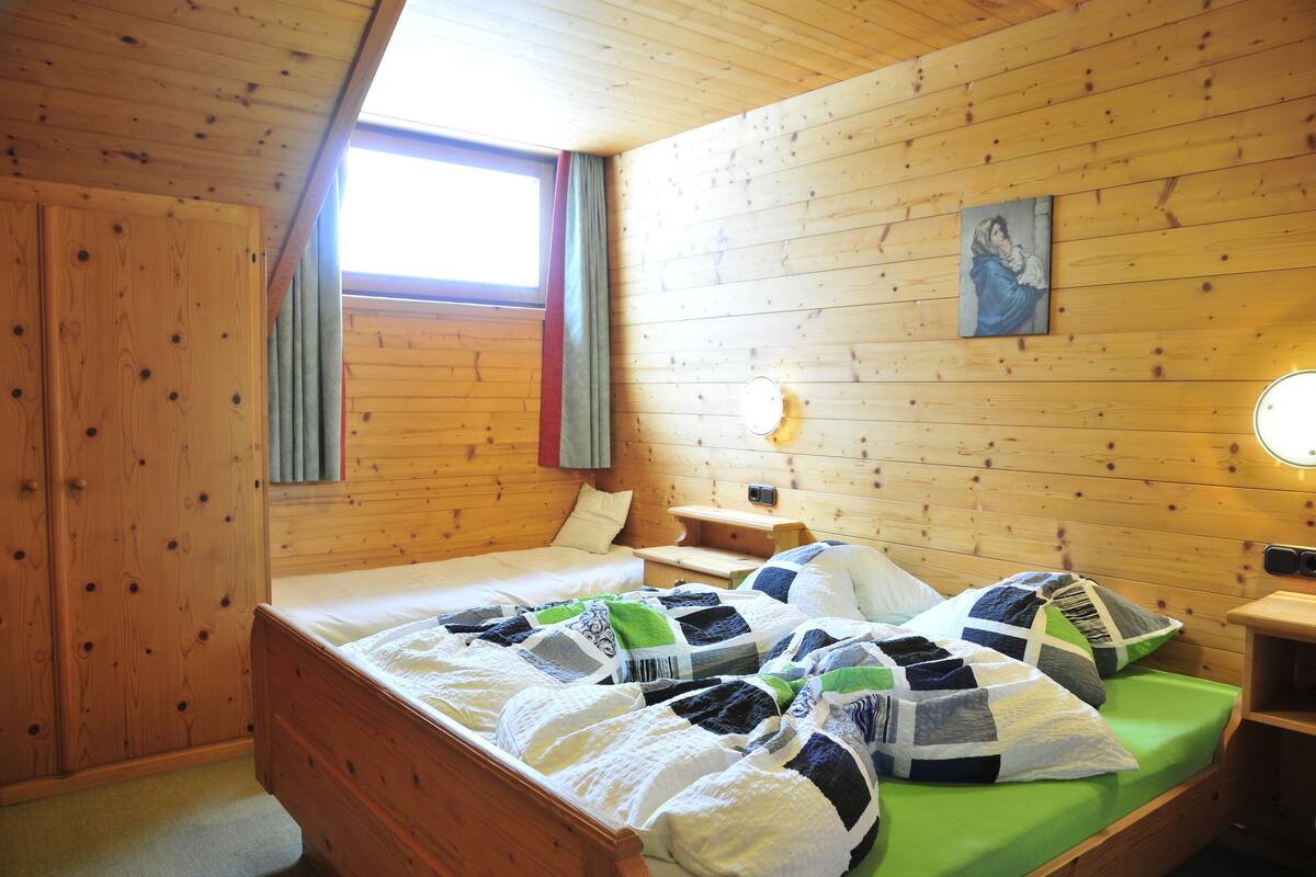 Quarto com painéis de madeira, uma cama, uma mesinha de cabeceira, uma janela com cortinas e um quadro na parede.