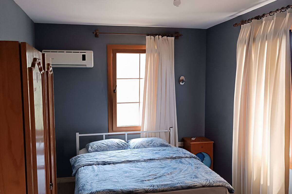 Quarto com cama, guarda-roupa de madeira, mesa de cabeceira e janela com cortinas.