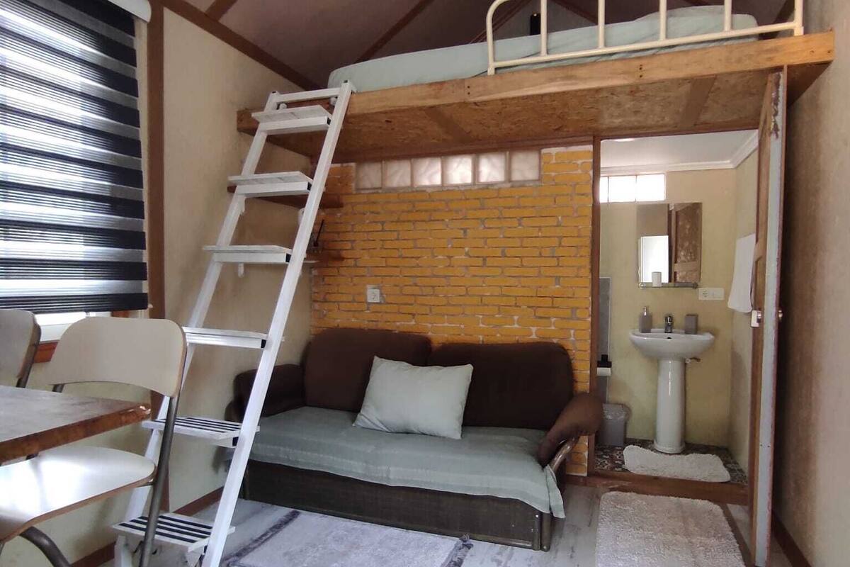 Quarto compacto com beliche, escrivaninha com cadeira, escada, sofá, banheiro pequeno com pia e janela com persianas.